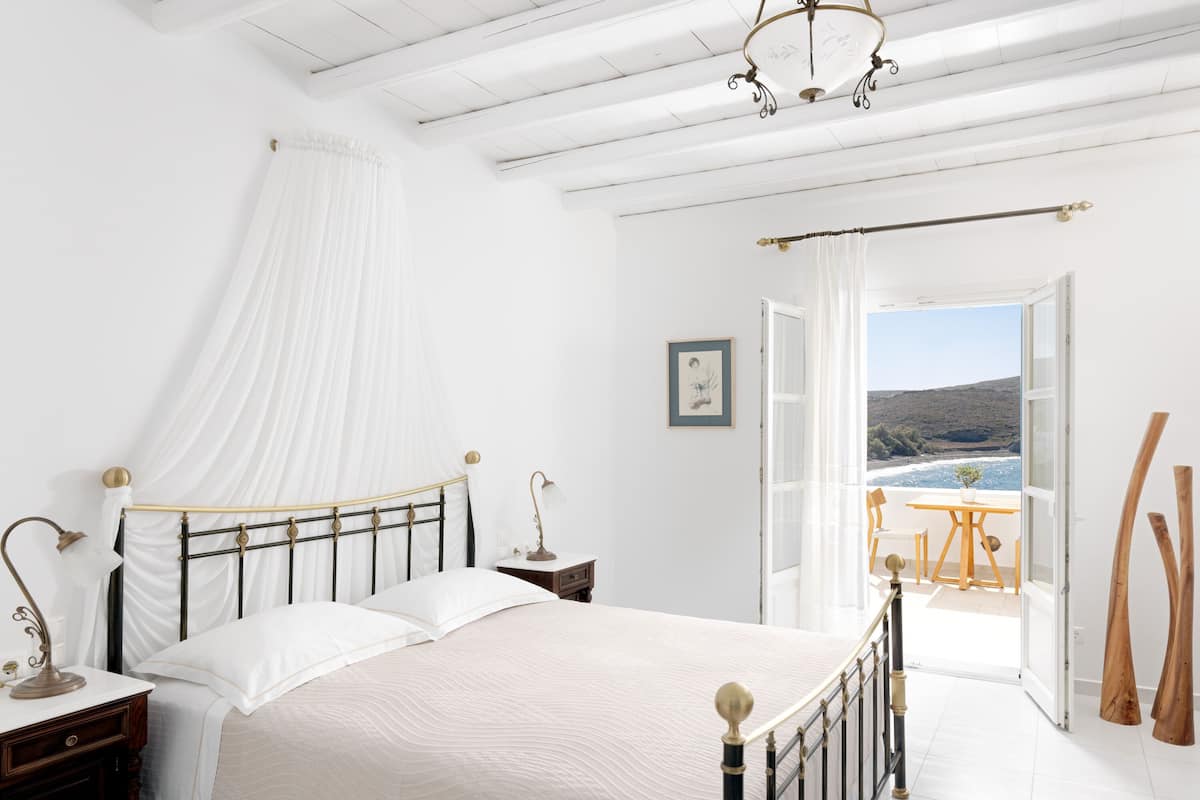 Quarto com cama, mesa de cabeceira, abajur e vista para o mar através de uma porta aberta.