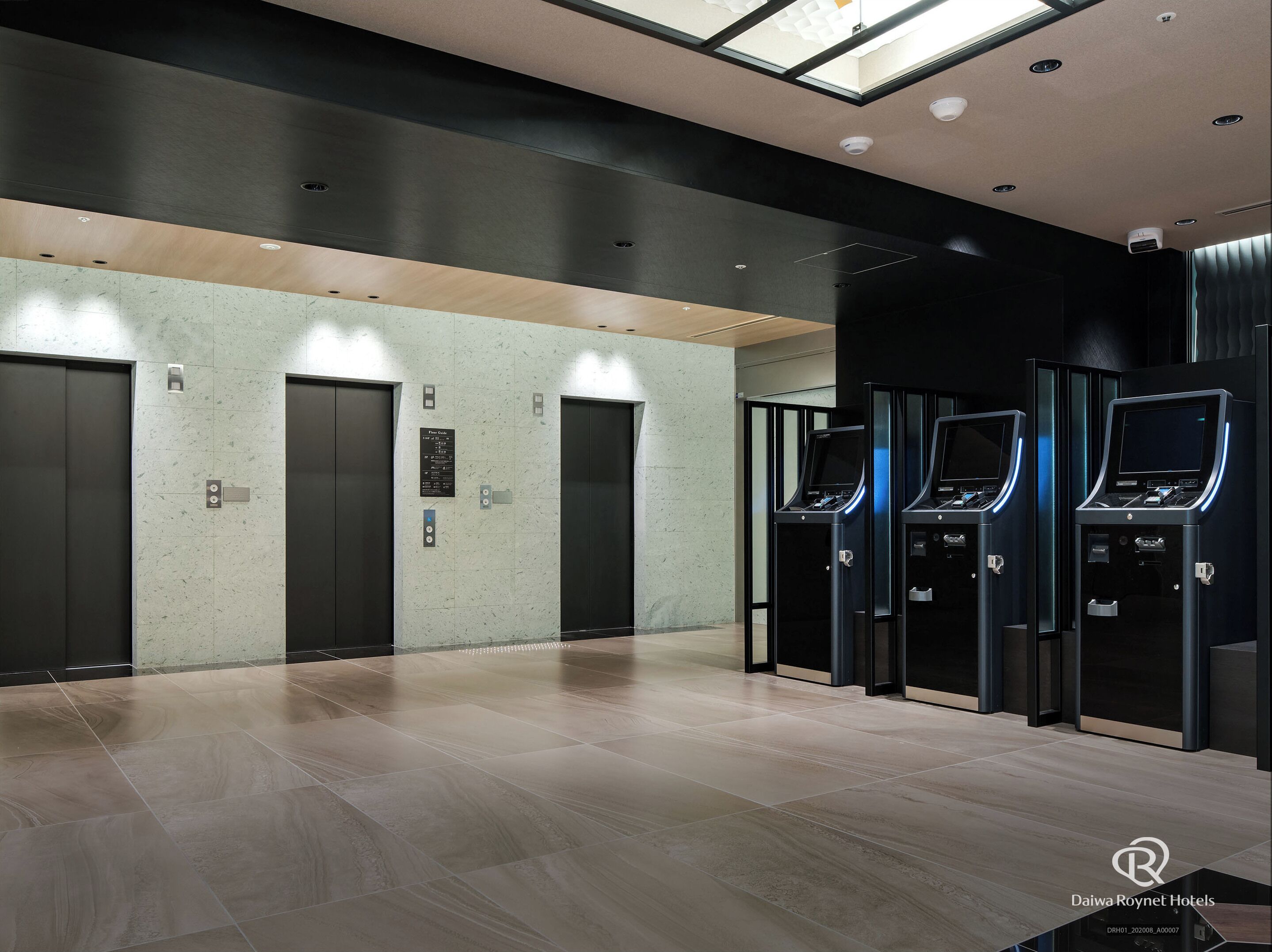 Área de elevadores com design moderno, incluindo um painel de informações digital e um quiosque de autoatendimento.