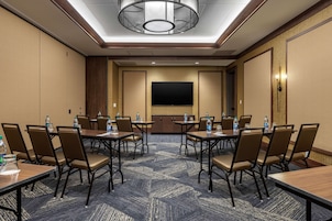 Sala de conferência com uma televisão de tela grande, várias mesas com cadeiras e garrafas de água em cada mesa.