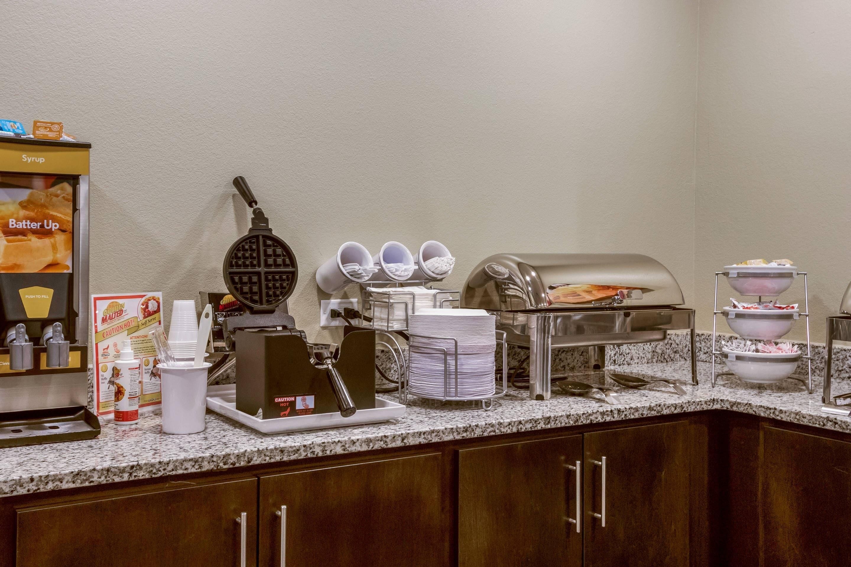Um balcão de café da manhã com máquina de waffles, cafeteira e pratos empilhados.