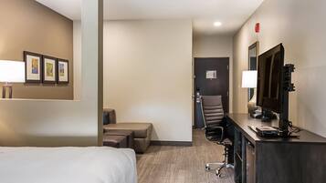 Quarto de hotel com uma cama, uma escrivaninha com computador, uma cadeira e um pequeno espaço para sentar.