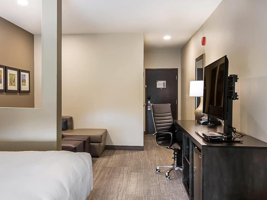 Quarto de hotel com uma cama, uma escrivaninha com computador, uma cadeira e um pequeno espaço para sentar.