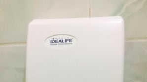 Um dosificador branco Idealife com dois botões identificados como 'Champô' e 'Gel de Banho' em frente a uma parede revestida a azulejos.