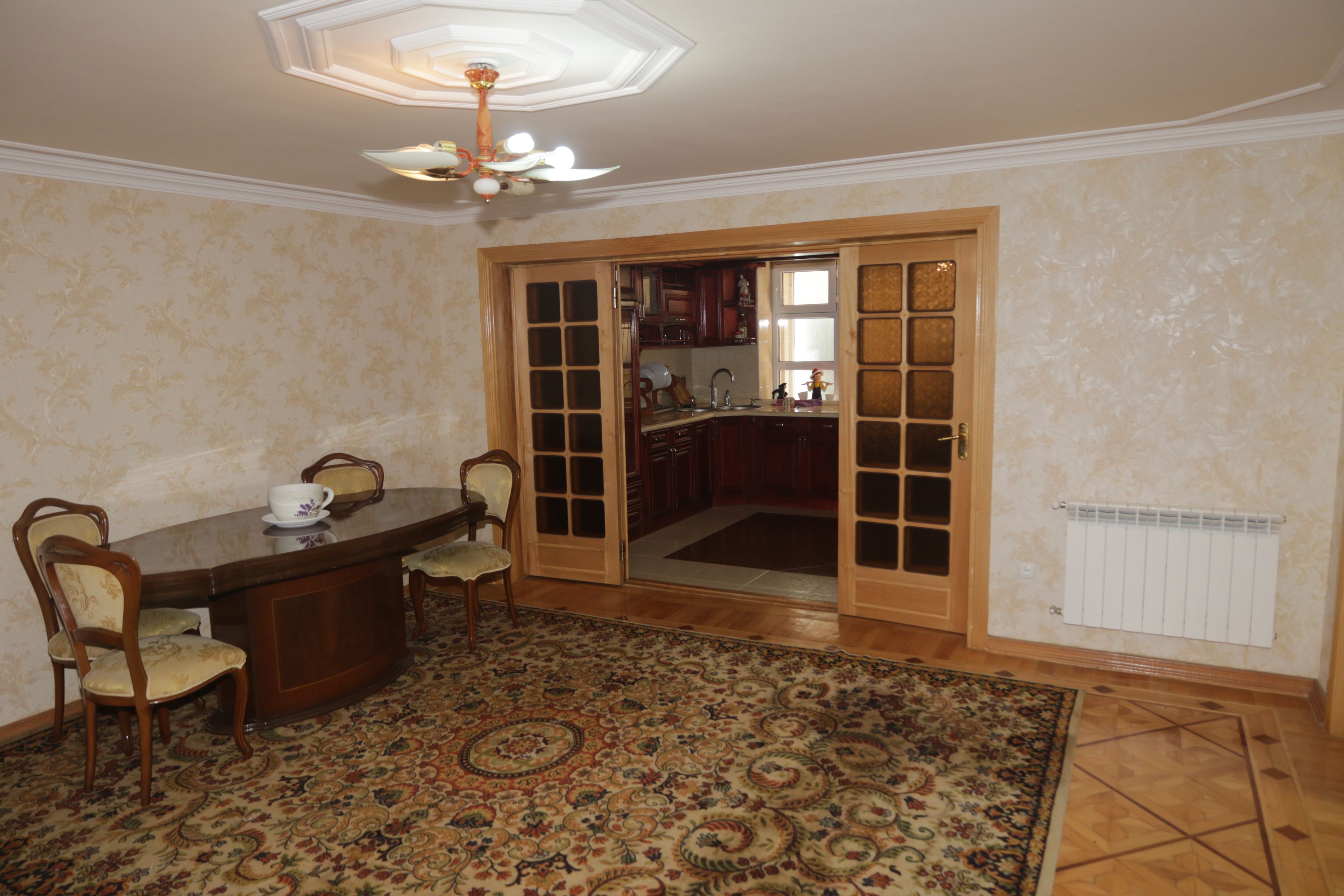 Uma sala de jantar com mesa e cadeiras de madeira, um tapete estampado e uma cozinha visível através de portas de vidro.