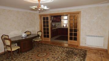 Uma sala de jantar com mesa e cadeiras de madeira, um tapete estampado e uma cozinha visível através de portas de vidro.