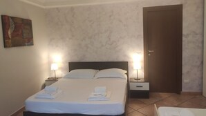 Uma cama bem arrumada com lençóis e toalhas brancas, duas luminárias de cabeceira, uma porta e um quadro na parede.