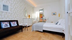 Quarto de hotel moderno com parede de padrão geométrico, piso em madeira com padrão espinha de peixe, cama branca, sofá marrom com almofadas de tema canino, escrivaninha branca pequena e mesa de cabeceira com luminária.
