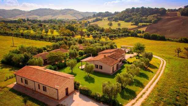 Vista aérea de uma propriedade rural com vários edifícios, rodeada por campos verdes e árvores, com uma paisagem montanhosa ao fundo.