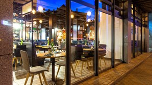 Um restaurante interno com mesas, cadeiras, janelas amplas e piso de tijolos.