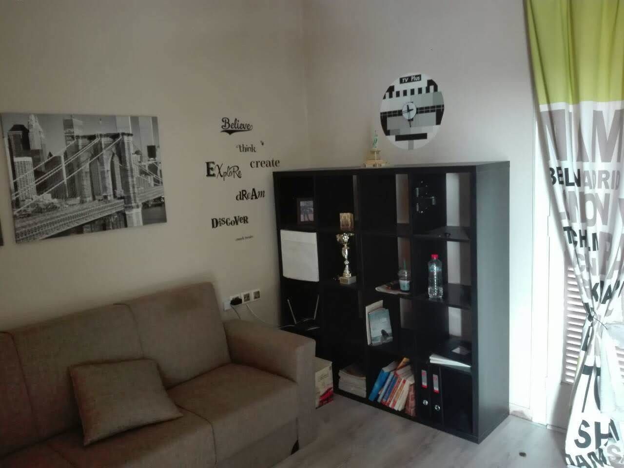 Uma sala de estar com um sofá, uma estante preta, um quadro na parede e uma cortina.