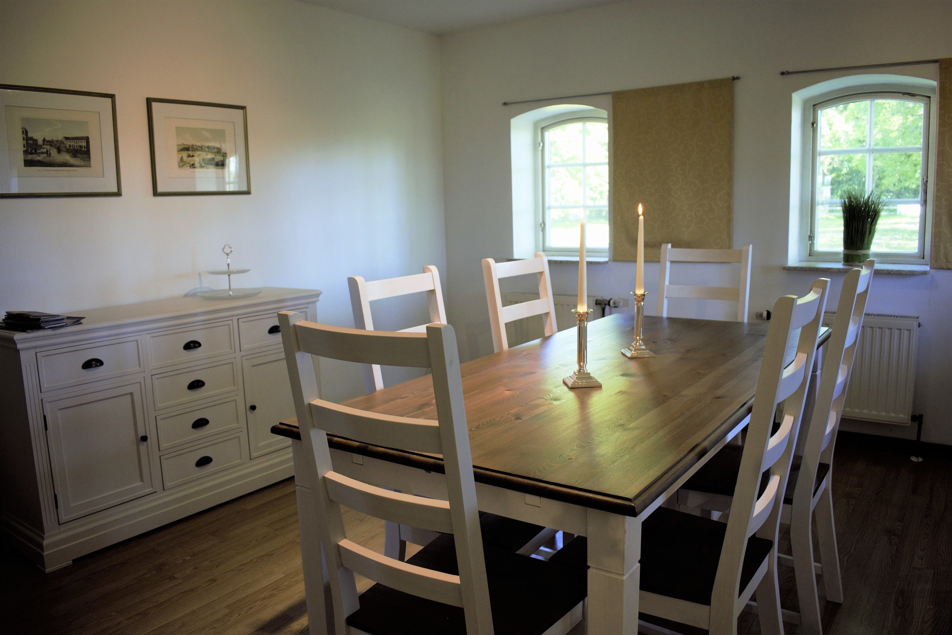 Uma sala de jantar com uma mesa de madeira, cadeiras brancas, um armário branco e duas molduras com quadros na parede.