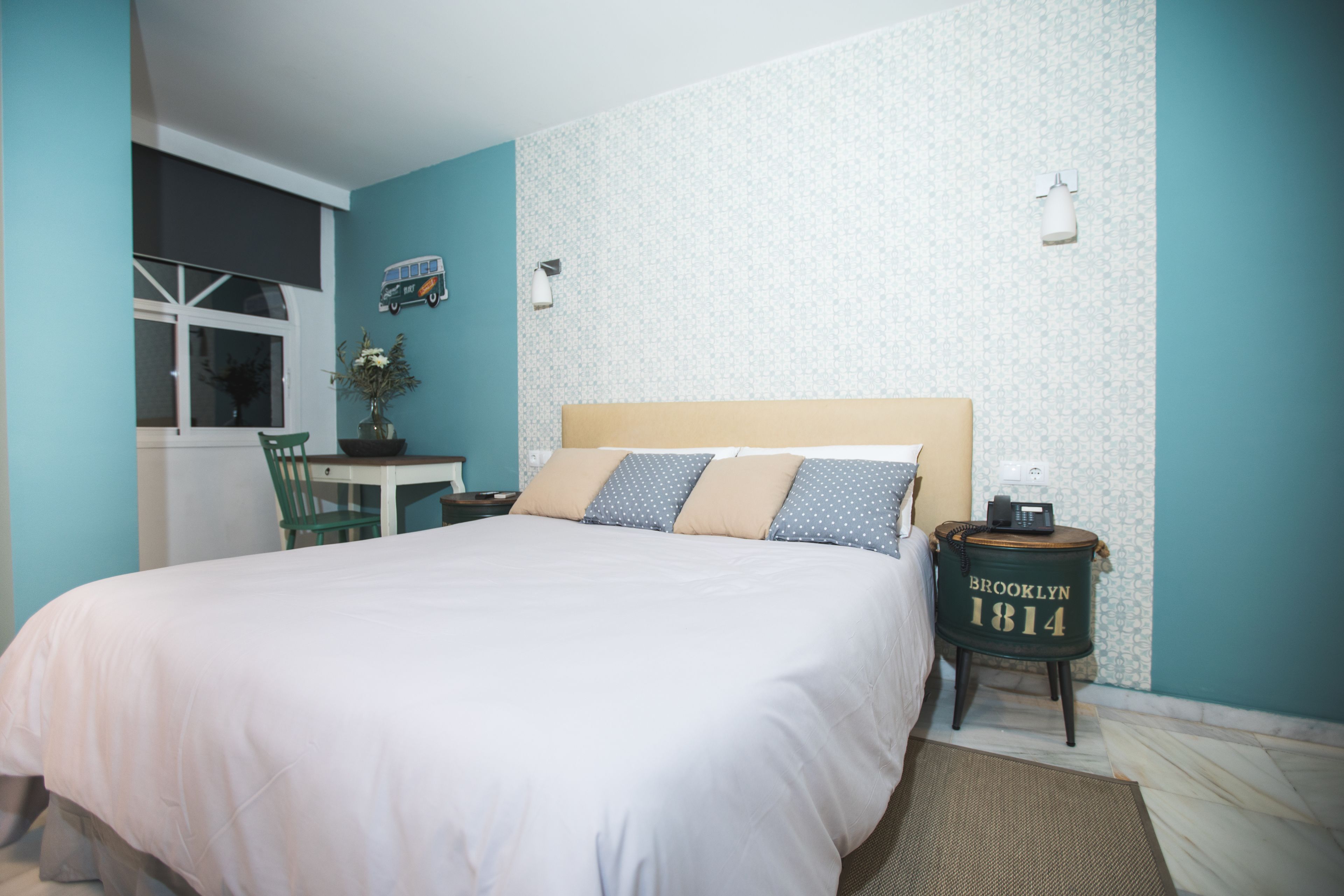 Quarto com cama grande, mesinha com cadeira, mesa de cabeceira com abajur e relógio de parede.