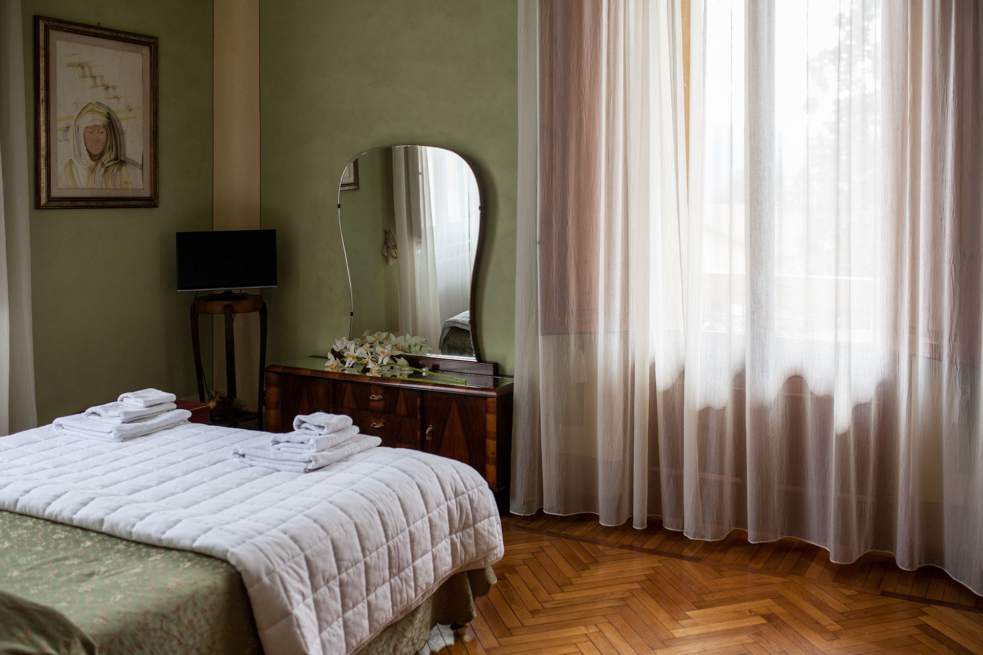Quarto de hotel com cama, penteadeira, espelho, televisão e janela com cortinas finas.