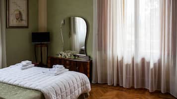 Quarto de hotel com cama, penteadeira, espelho, televisão e janela com cortinas finas.
