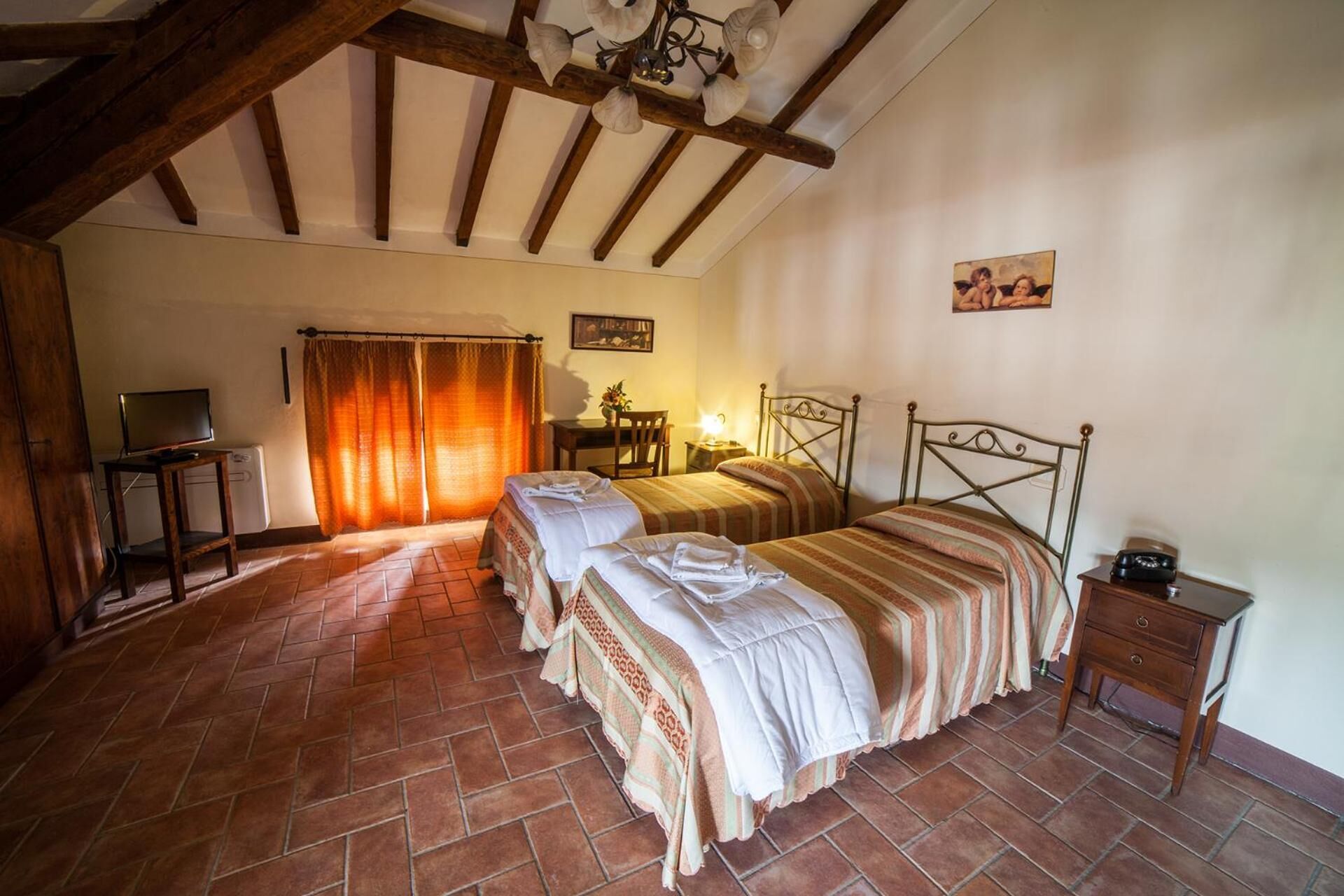 Quarto com duas camas, uma escrivaninha com computador, uma cadeira e uma mesinha de cabeceira com abajur.