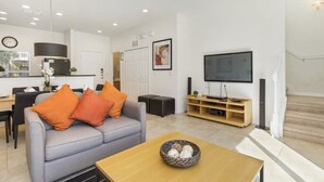 Uma sala de estar moderna com um sofá cinzento, almofadas laranja, uma mesa de centro de madeira, uma televisão de tela plana e uma escada que leva a outro andar.