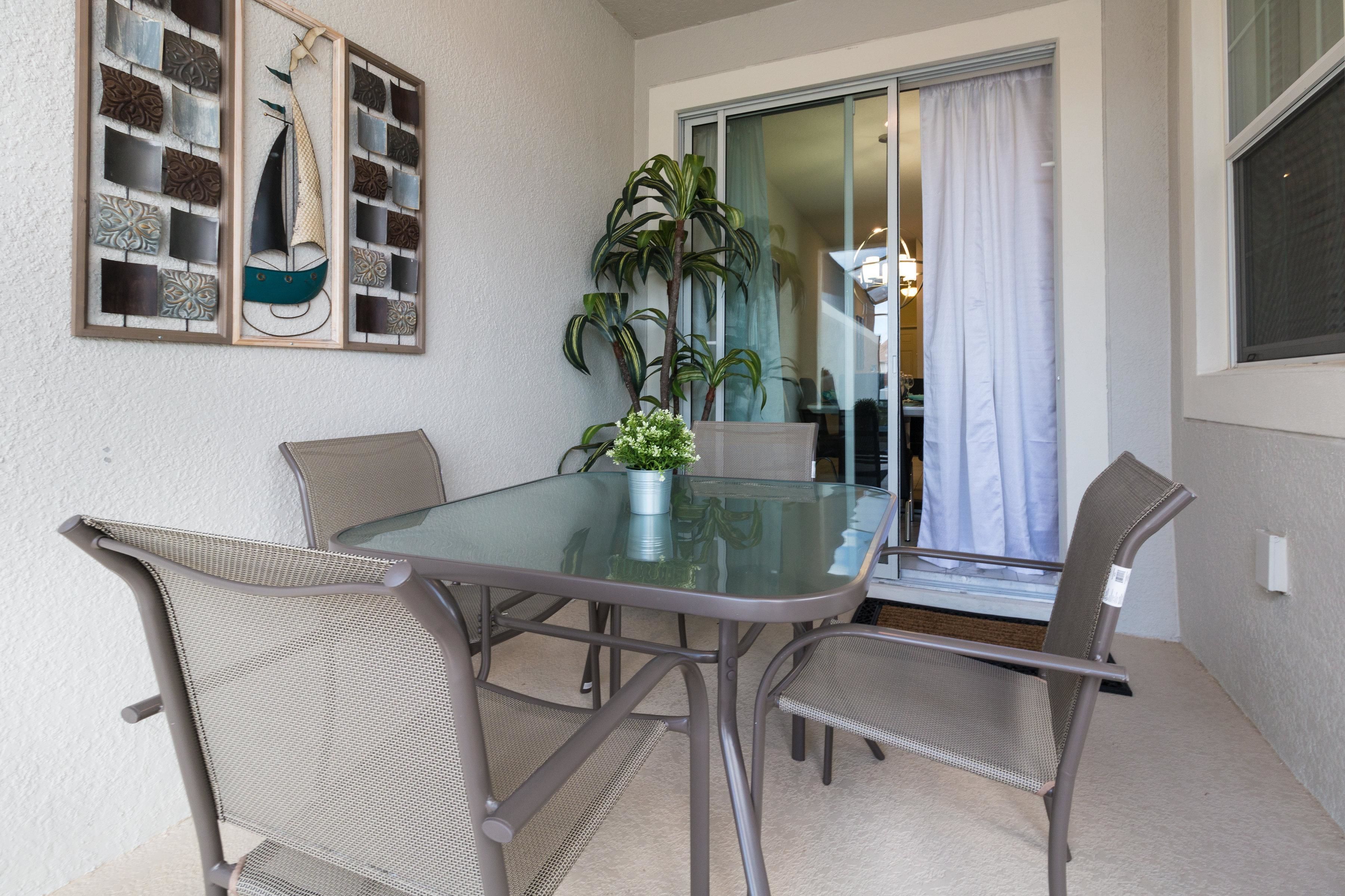 Uma mesa de jantar com tampo de vidro, quatro cadeiras, um vaso com planta e um quadro decorativo na parede.