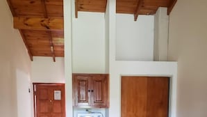 Quarto com uma cama, micro-ondas, armários de madeira, uma porta e uma mesa de cabeceira com abajur.