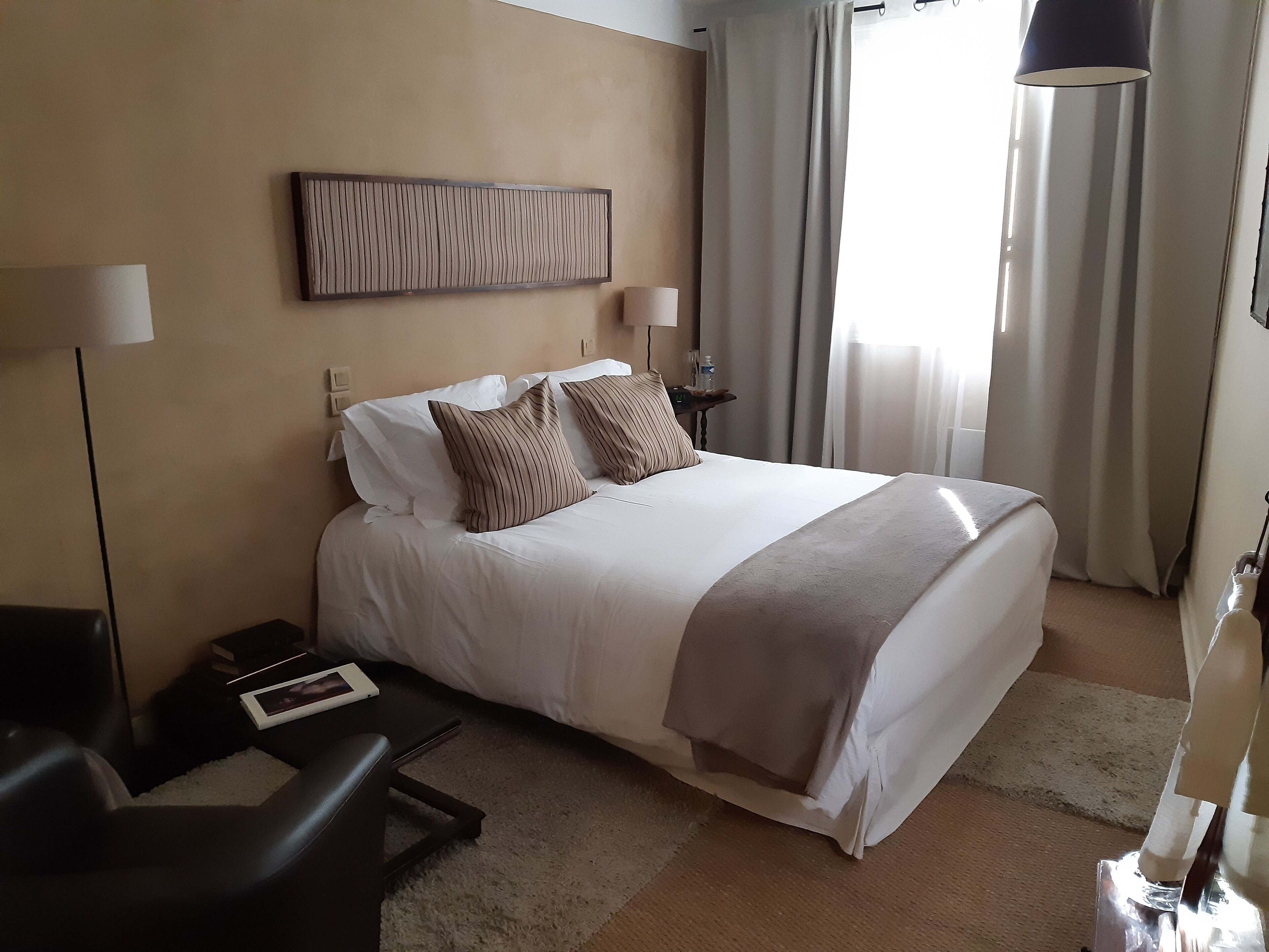 Quarto de hotel com uma cama grande, duas luminárias de cabeceira, uma obra de arte na parede, uma mesa com cadeira e uma janela com cortinas.