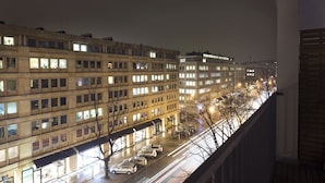Um edifício com vários andares, janelas iluminadas, área de estacionamento com carros e uma rua com semáforos.