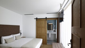 Quarto de hotel com uma cama grande, porta de madeira, banheiro com espelho e um banco.