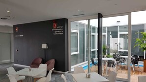 Um espaço moderno e interno com mesas e cadeiras brancas, uma parede preta com placas de sinalização e grandes portas de vidro que dão para uma área externa com piscina.