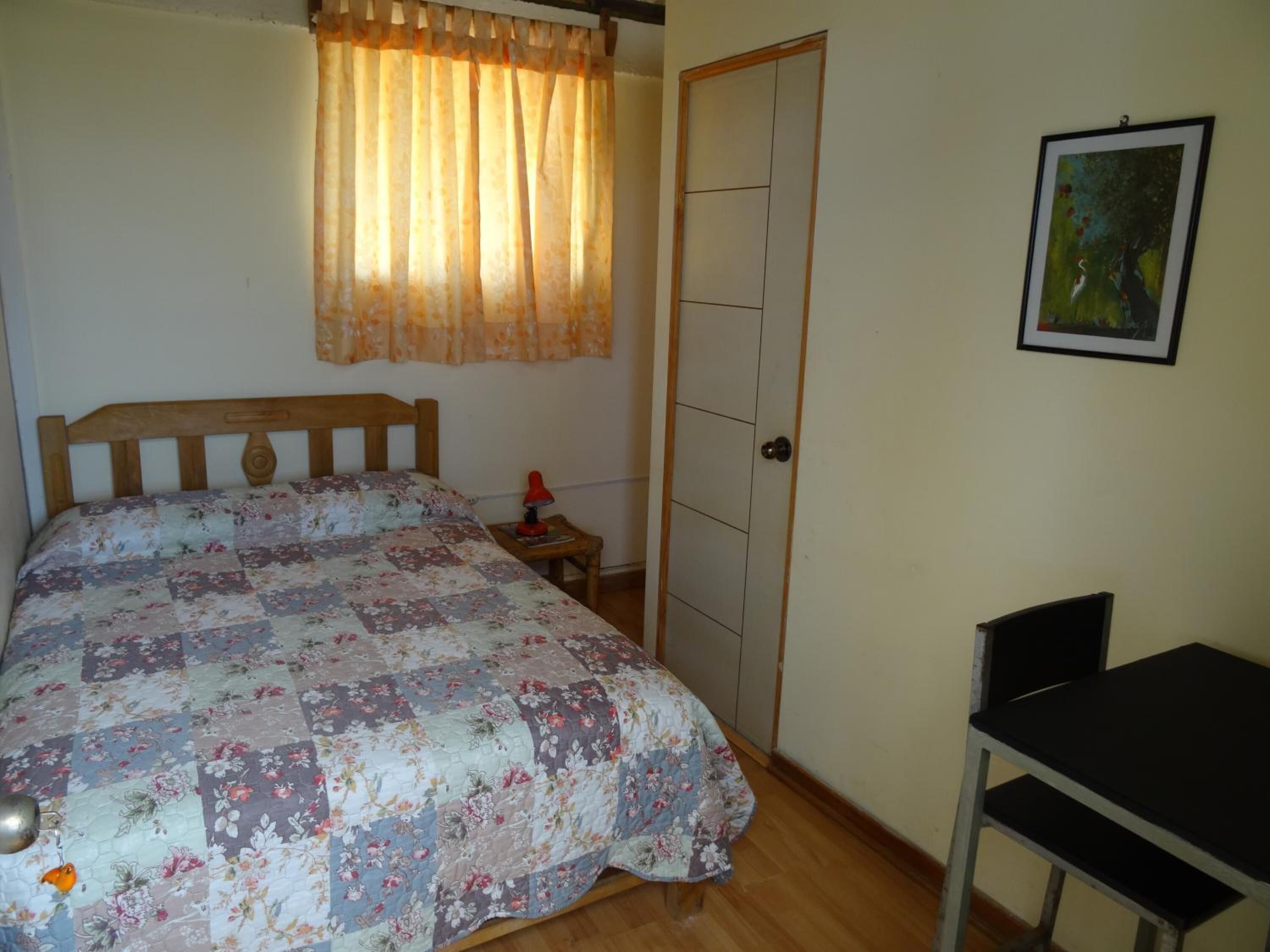 Quarto com cama de madeira, mesa de cabeceira pequena com abajur, porta e quadro pendurado na parede.