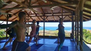 Um grupo de pessoas praticando yoga em tapetes sob um telhado de madeira, com vista para o oceano.