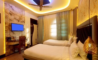 Quarto de hotel com uma cama, escrivaninha com televisão, ventoinha no teto e janela com cortinas.
