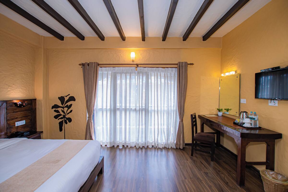 Quarto de hotel com cama, escrivaninha, cadeira, televisão e uma janela grande com cortinas.