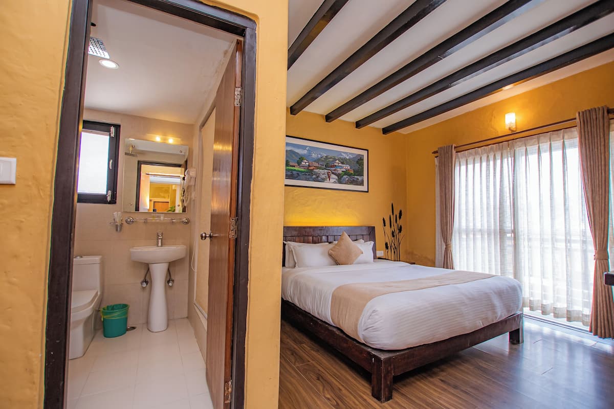 Quarto de hotel com cama de madeira, banheiro com pia e vaso sanitário, e uma janela grande com cortinas.