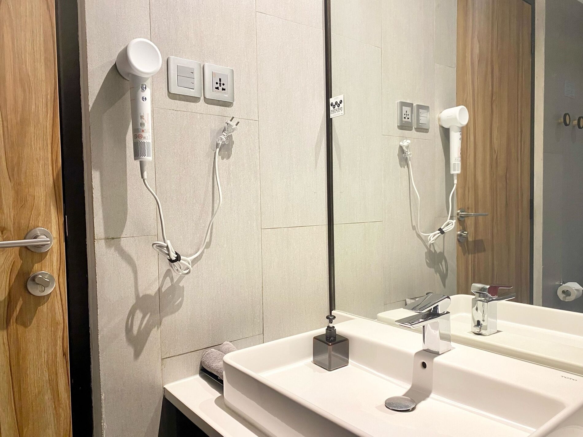 Casa de banho com lavatório branco, espelho, toalheiro e dois secadores de cabelo fixados na parede.