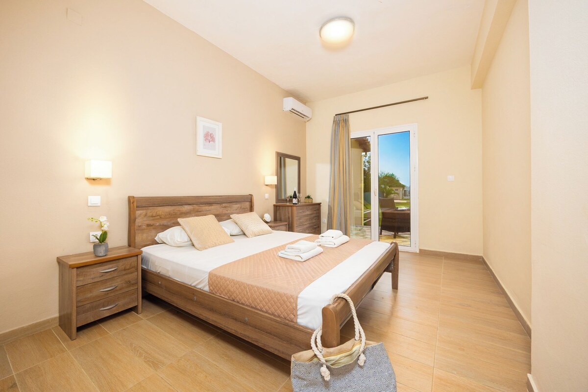 Quarto de hotel com cama de madeira, mesa de cabeceira, cômoda, espelho e porta de vidro corrediça para a varanda.
