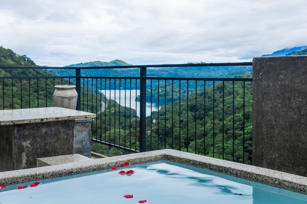 Uma piscina com nenúfares, um banco de pedra e vista para um lago rodeado por colinas.