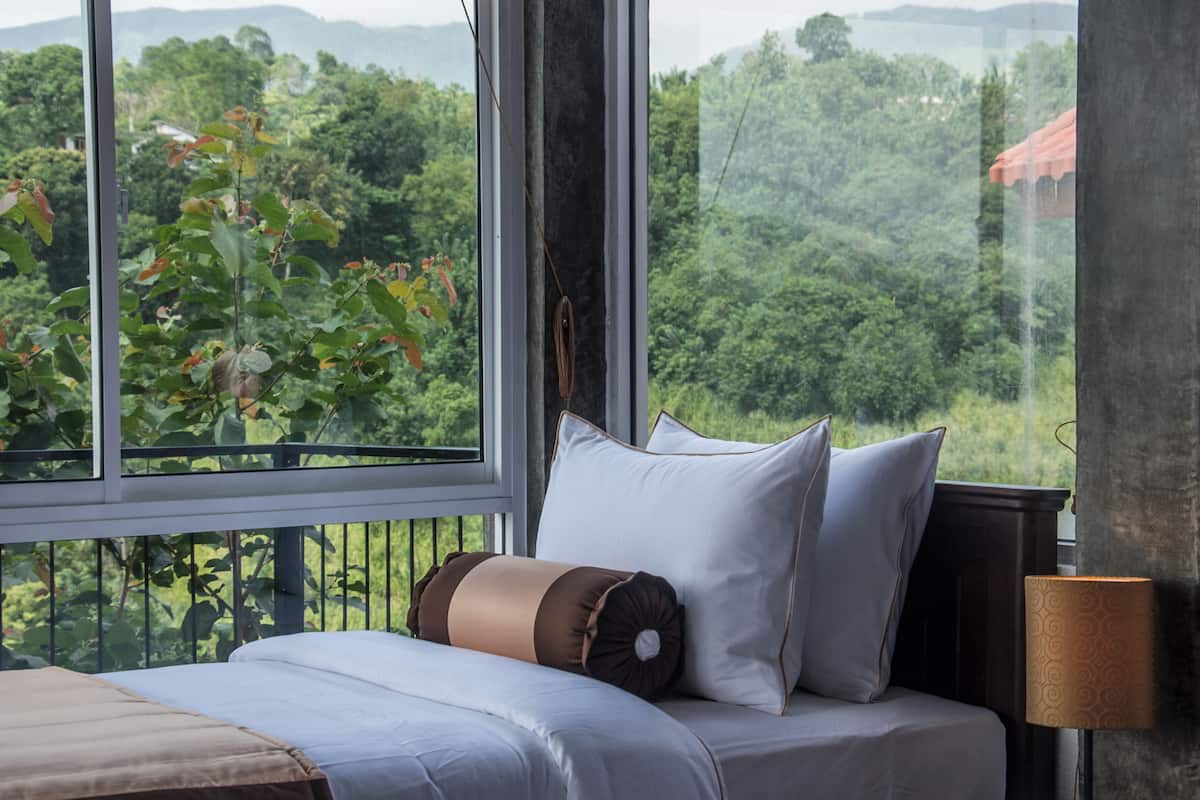 Quarto com cama, almofadas e janela com vista para uma área arborizada.