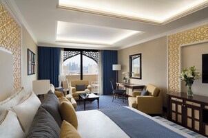 Quarto de hotel com uma cama grande, um sofá, uma escrivaninha e uma cadeira. Vê-se a cidade pelas janelas.