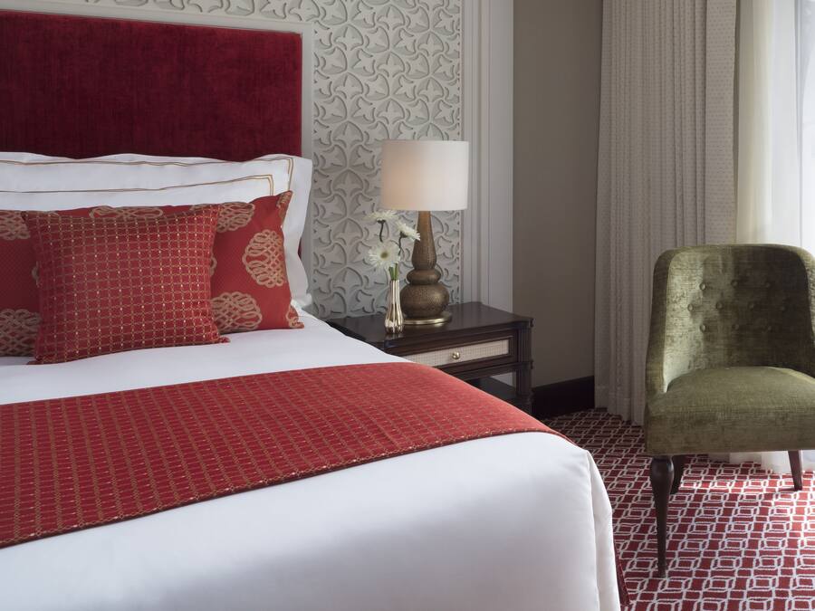 Quarto de hotel com uma cama grande, uma mesa de cabeceira com abajur, uma poltrona verde e papel de parede estampado.