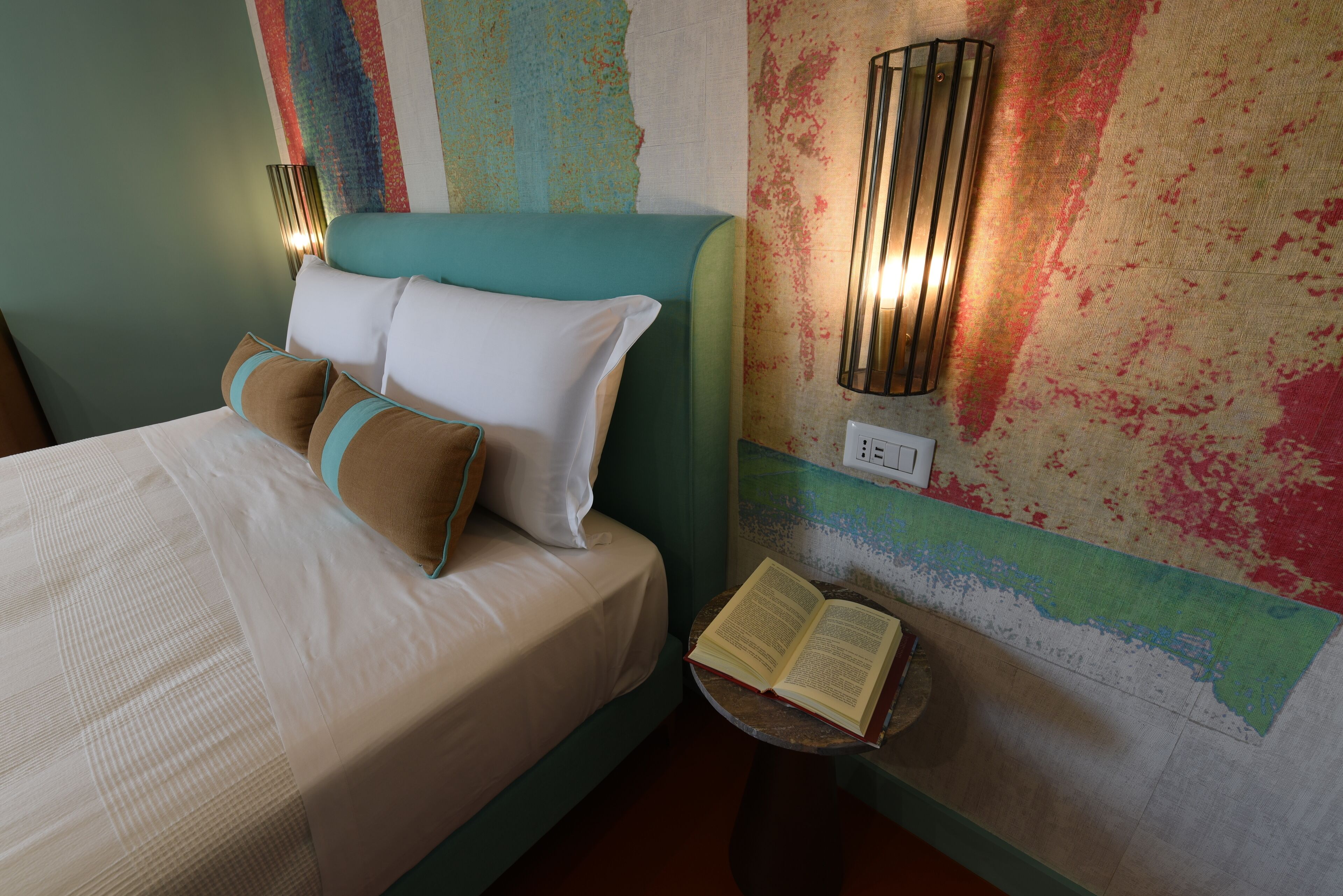 Uma cama bem arrumada com almofadas, uma mesa de cabeceira com um livro aberto e uma luminária fixada na parede.