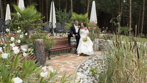 Um casal sentado num banco num jardim com mobiliário de exterior e plantas tropicais.