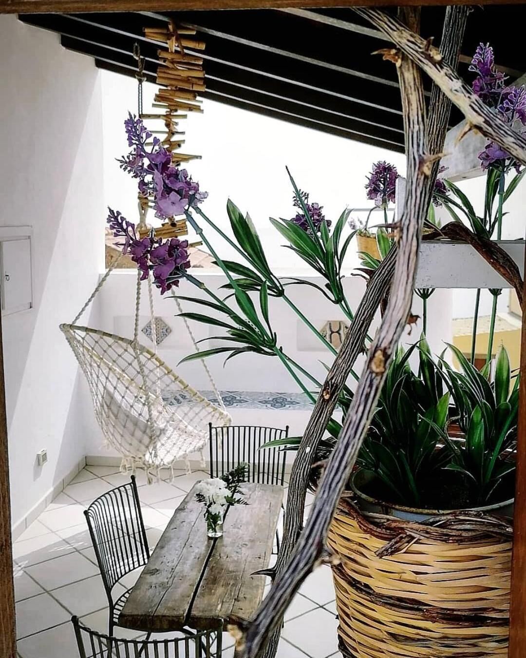 Uma área de refeições com uma mesa e cadeiras de madeira, cercada por plantas em vasos e flores roxas pendentes.