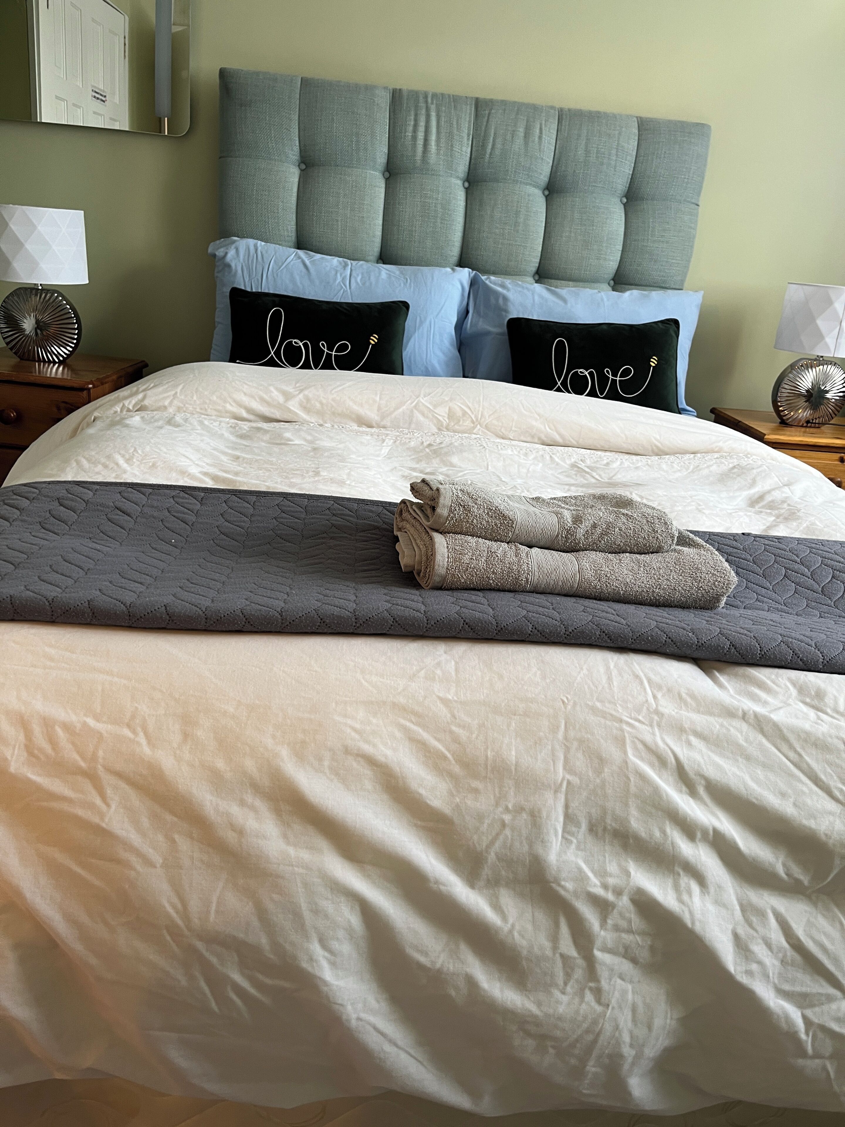 Cama bem arrumada com colcha cinza, duas toalhas dobradas e almofadas com a palavra 'love'.