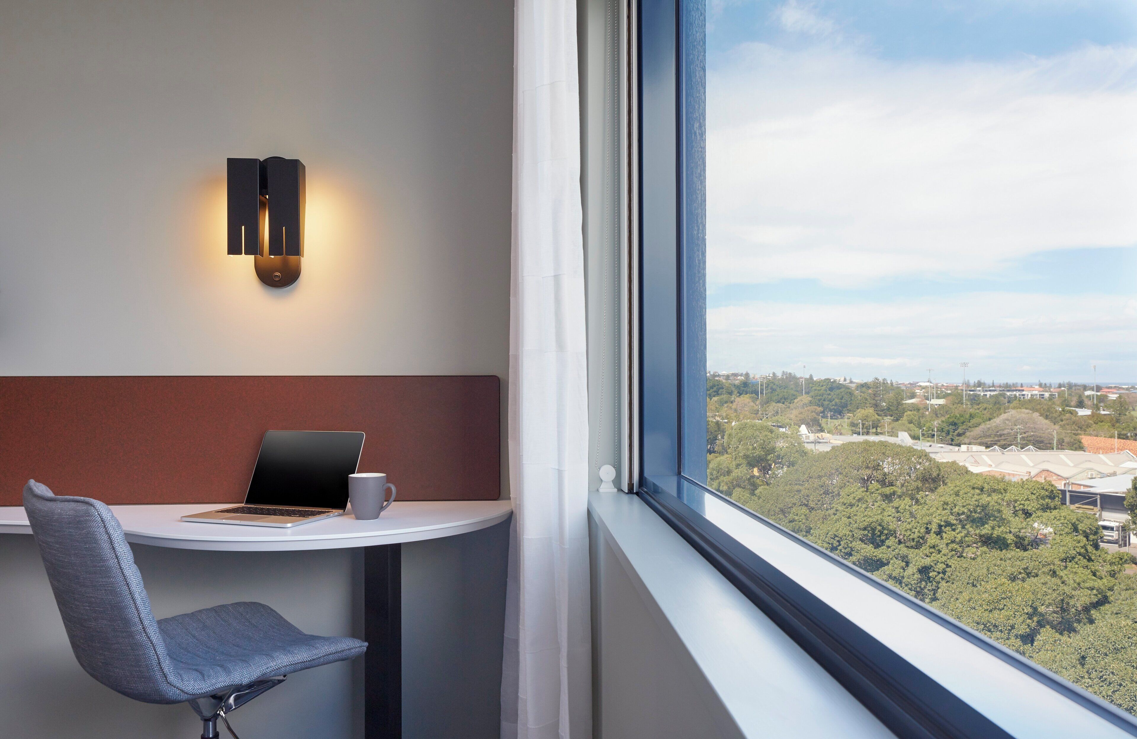 Quarto de hotel moderno com escrivaninha, cadeira e vista para a paisagem urbana através de uma janela grande.