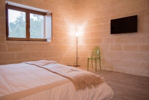 Quarto com cama, janela, luminária de chão, cadeira e televisão fixada na parede.