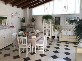 Uma sala de jantar com uma mesa posta para quatro pessoas, rodeada por cadeiras brancas. Há um sofá branco e uma grande planta em vaso no canto.