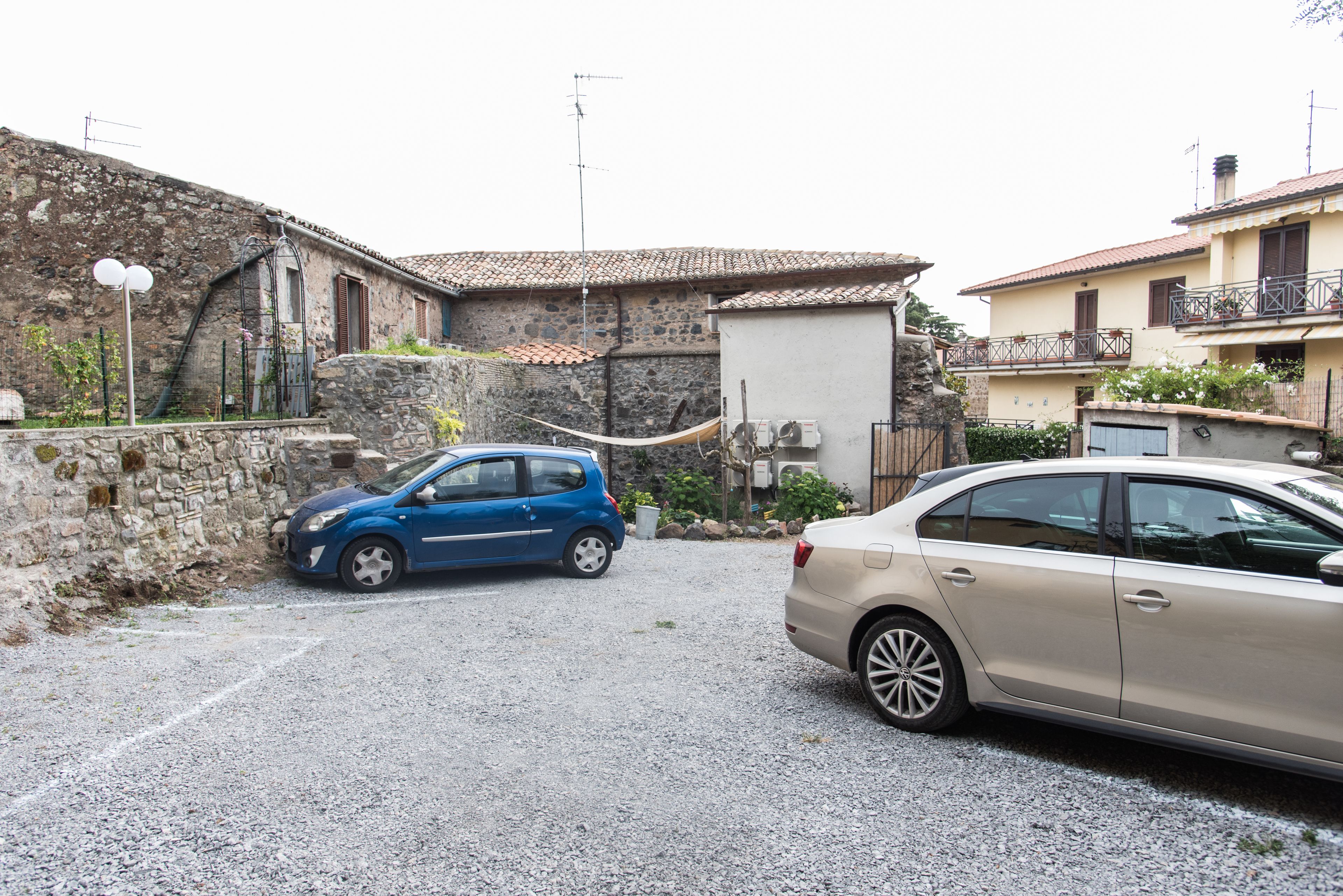 Rua com dois carros estacionados, um edifício de pedra e uma casa com telhado de cerâmica.