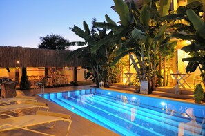 Uma piscina com espreguiçadeiras, cercada por plantas tropicais e uma estrutura com telhado de palha.
