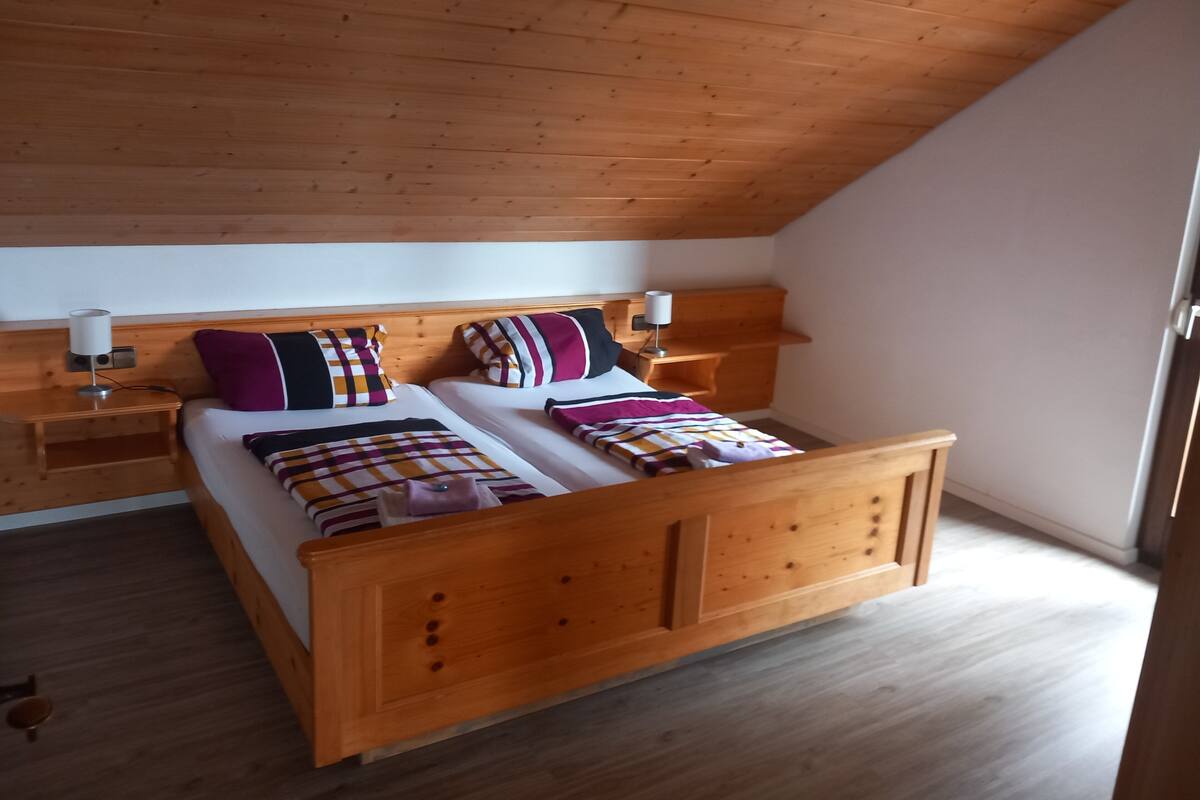 Uma cama de madeira com duas mesas de cabeceira, cada uma com uma luminária, em um quarto com teto inclinado.