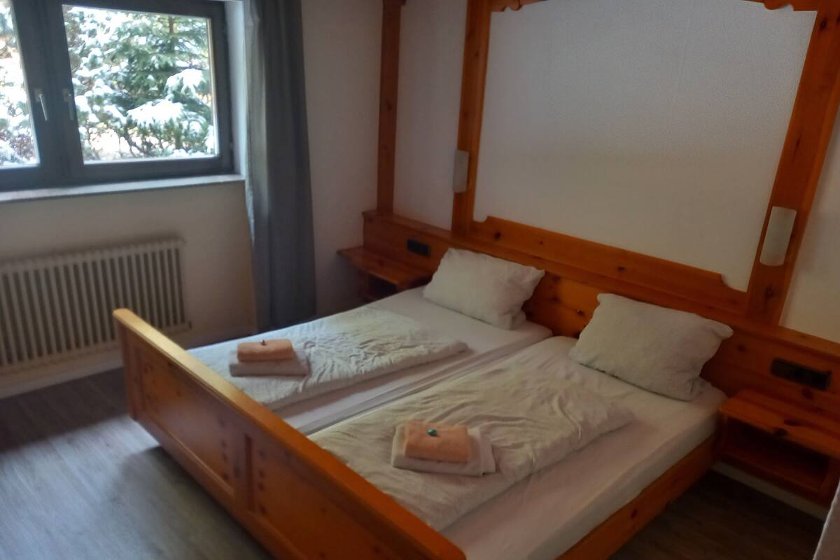 Uma cama de madeira com duas almofadas e um cobertor, uma mesa de cabeceira, uma janela com vista para árvores e um radiador.