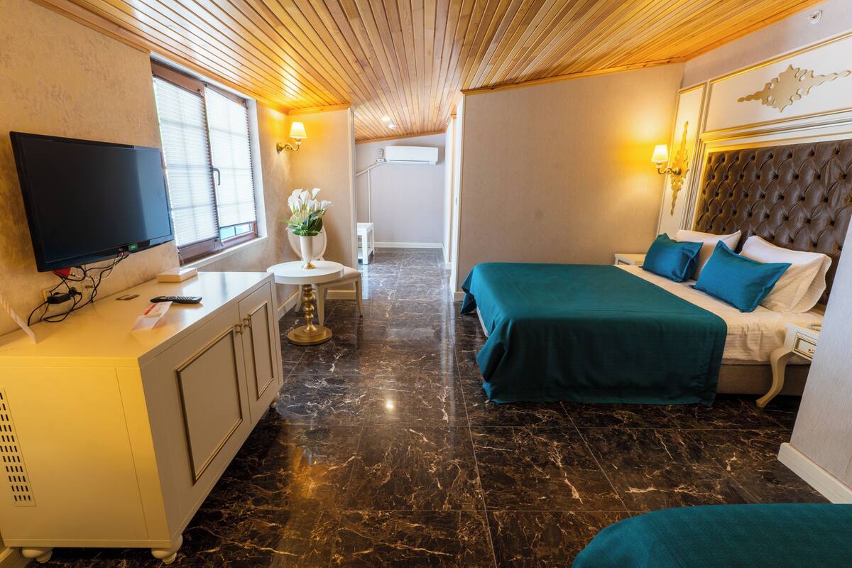 Quarto de hotel com televisão, escrivaninha com abajur, cama com roupa de cama azul-esverdeada e piso de mármore.