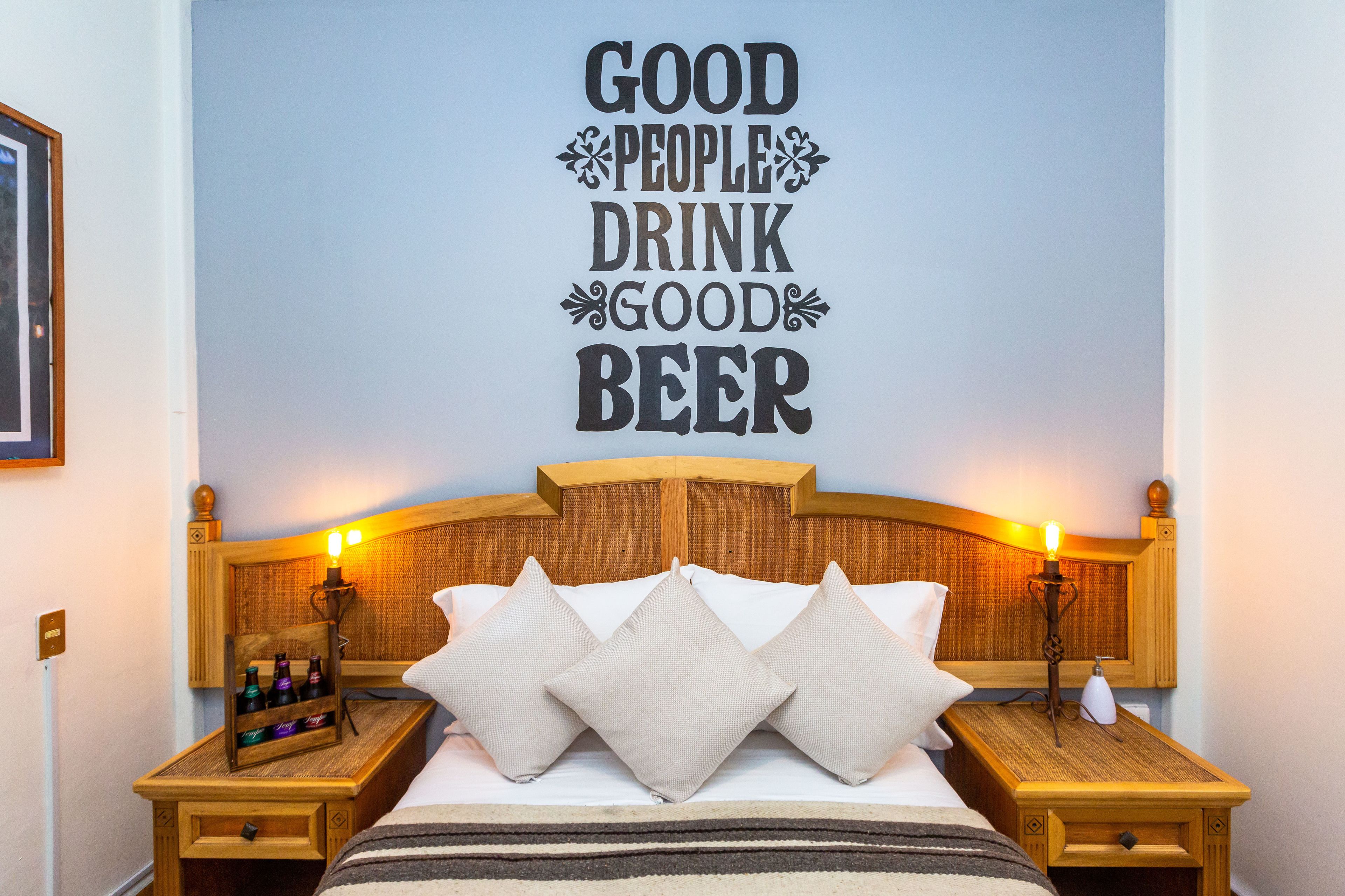 Quarto de hotel com cama, almofadas, mesa de cabeceira com suporte para garrafa e uma parede com mensagem inspiradora.